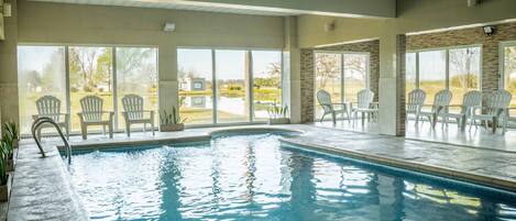 Piscina interior com paredes de vidro transparente, rodeada por cadeiras brancas, e vista para área ajardinada externa.