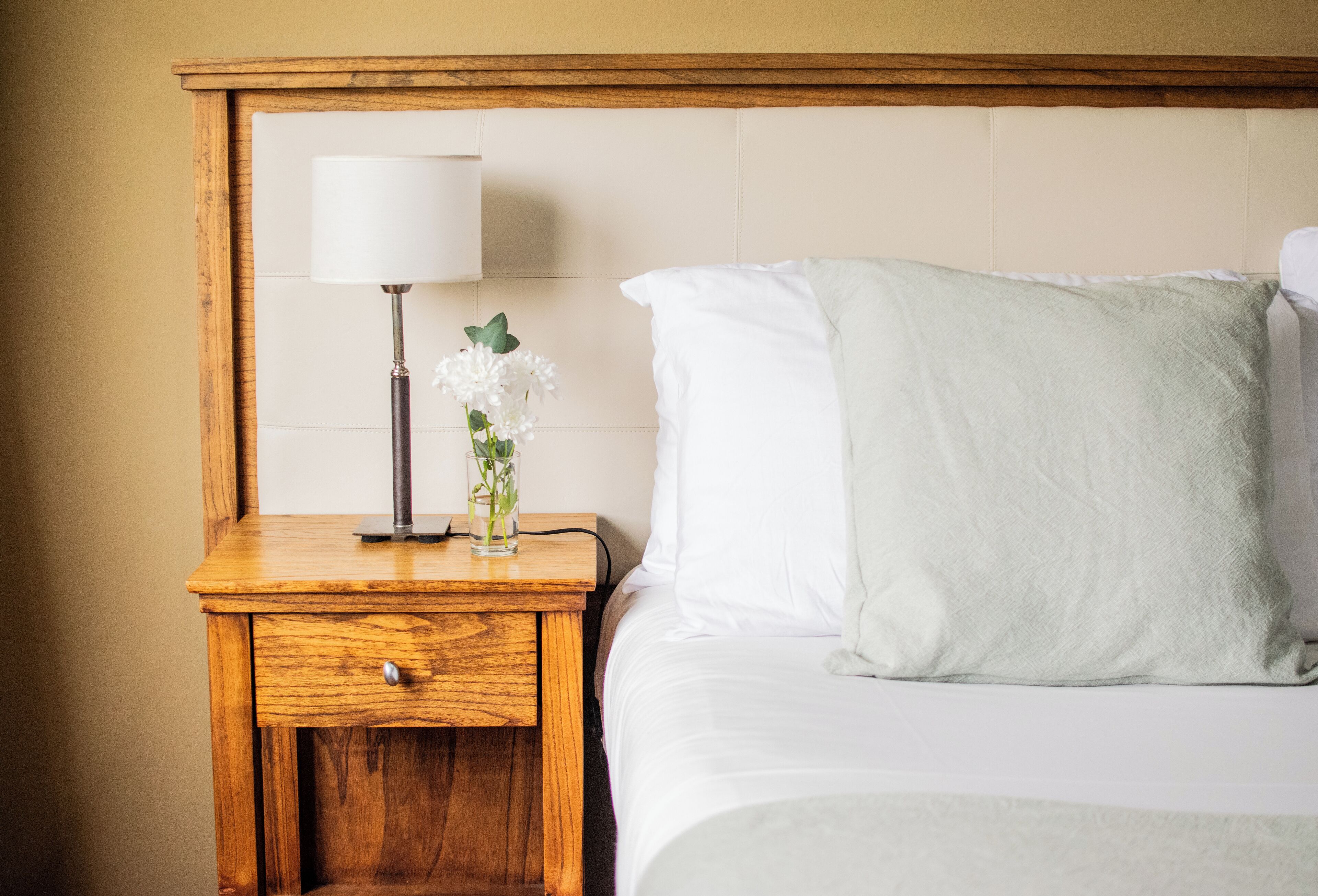 Cama bem arrumada com lençóis brancos e almofada verde-claro, mesa de cabeceira de madeira com abajur e vaso de vidro com flores, e uma parede lisa ao fundo.
