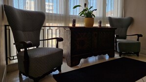 Quarto com duas poltronas, um aparador de madeira, um vaso com planta e vista através de cortinas leves.