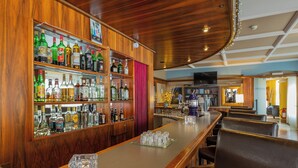 Uma área de bar com prateleiras de madeira repletas de garrafas de bebidas, um balcão com banquetas e uma televisão fixada na parede.