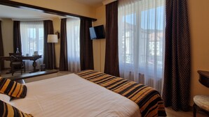 Quarto de hotel com uma cama grande, uma mesa com cadeira, uma televisão na parede e uma janela com cortinas.