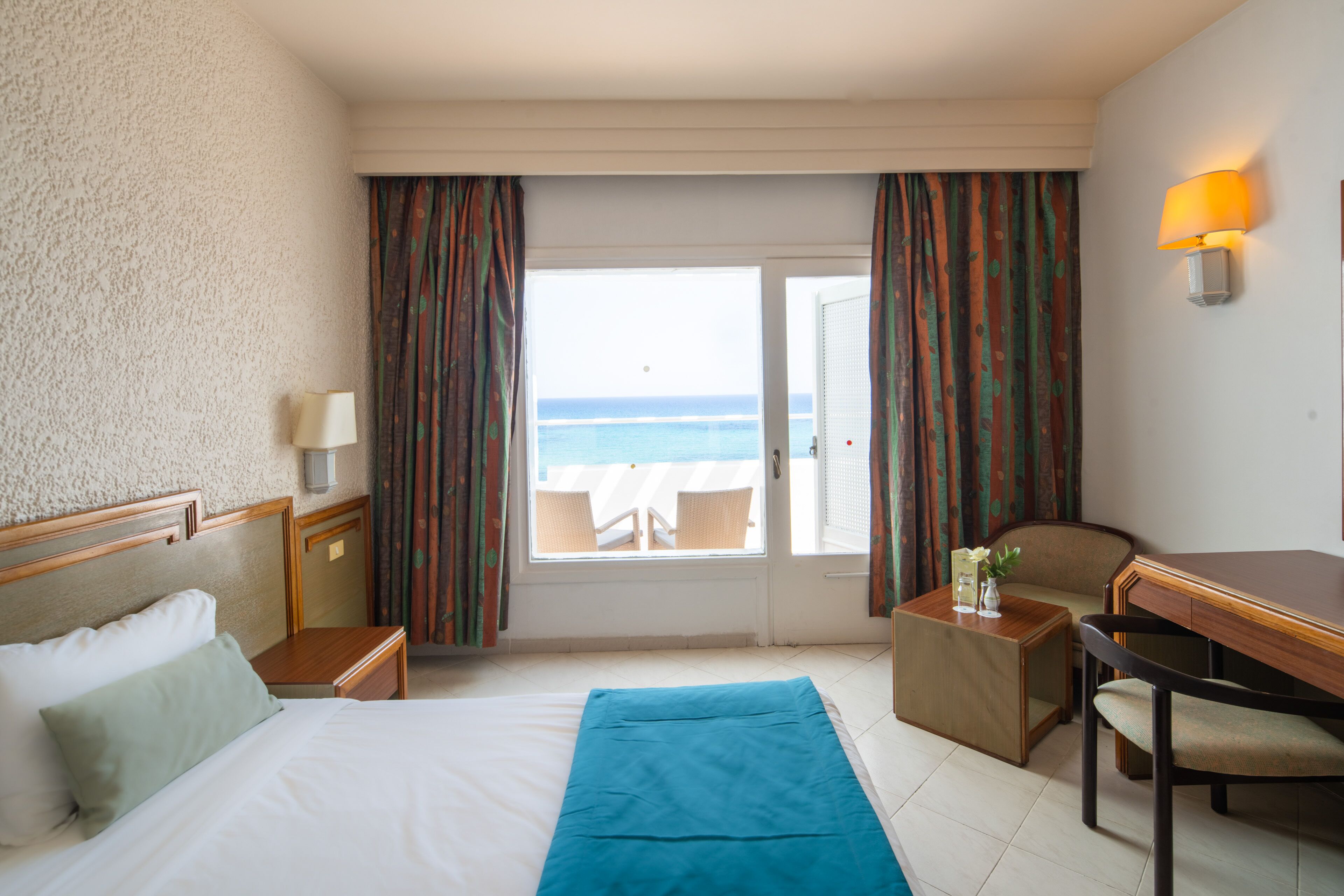 Quarto de hotel com cama, escrivaninha, cadeira, mesa de cabeceira e vista para o mar através de amplas janelas.