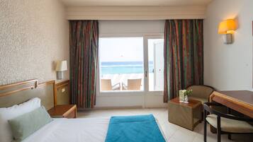 Quarto de hotel com cama, escrivaninha, cadeira, mesa de cabeceira e vista para o mar através de amplas janelas.