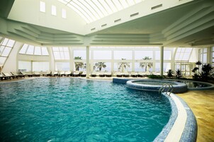 Piscina interior com vista clara para palmeiras e praia através de amplas janelas.