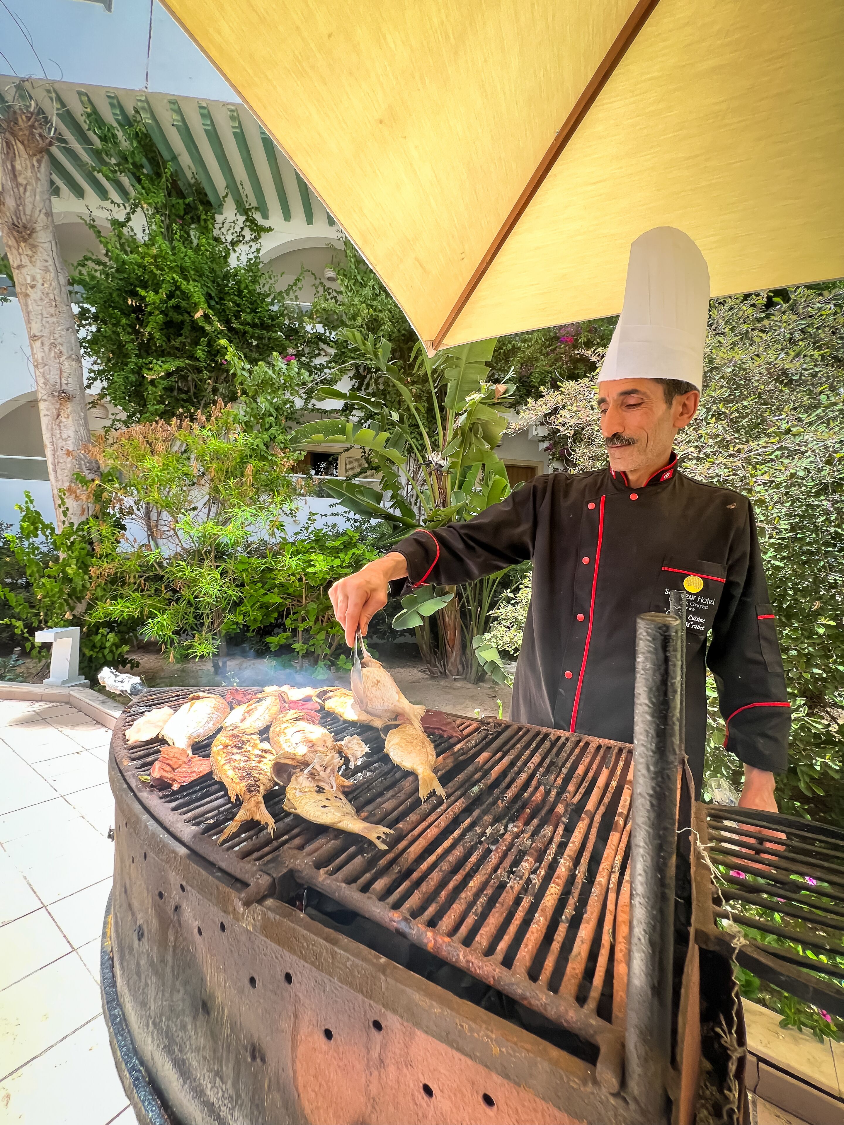 Um cozinheiro grelha diferentes carnes ao ar livre, com uma churrasqueira grande e um toldo acima.