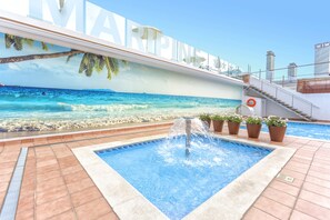 Uma piscina com uma fonte, rodeada por um deck ladrilhado e plantas em vasos.