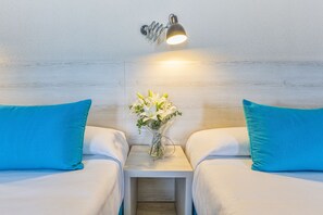 Quarto de hotel com duas camas, cada uma com travesseiro azul e roupa de cama branca. Uma pequena mesa de cabeceira com um vaso de flores brancas está entre as camas.