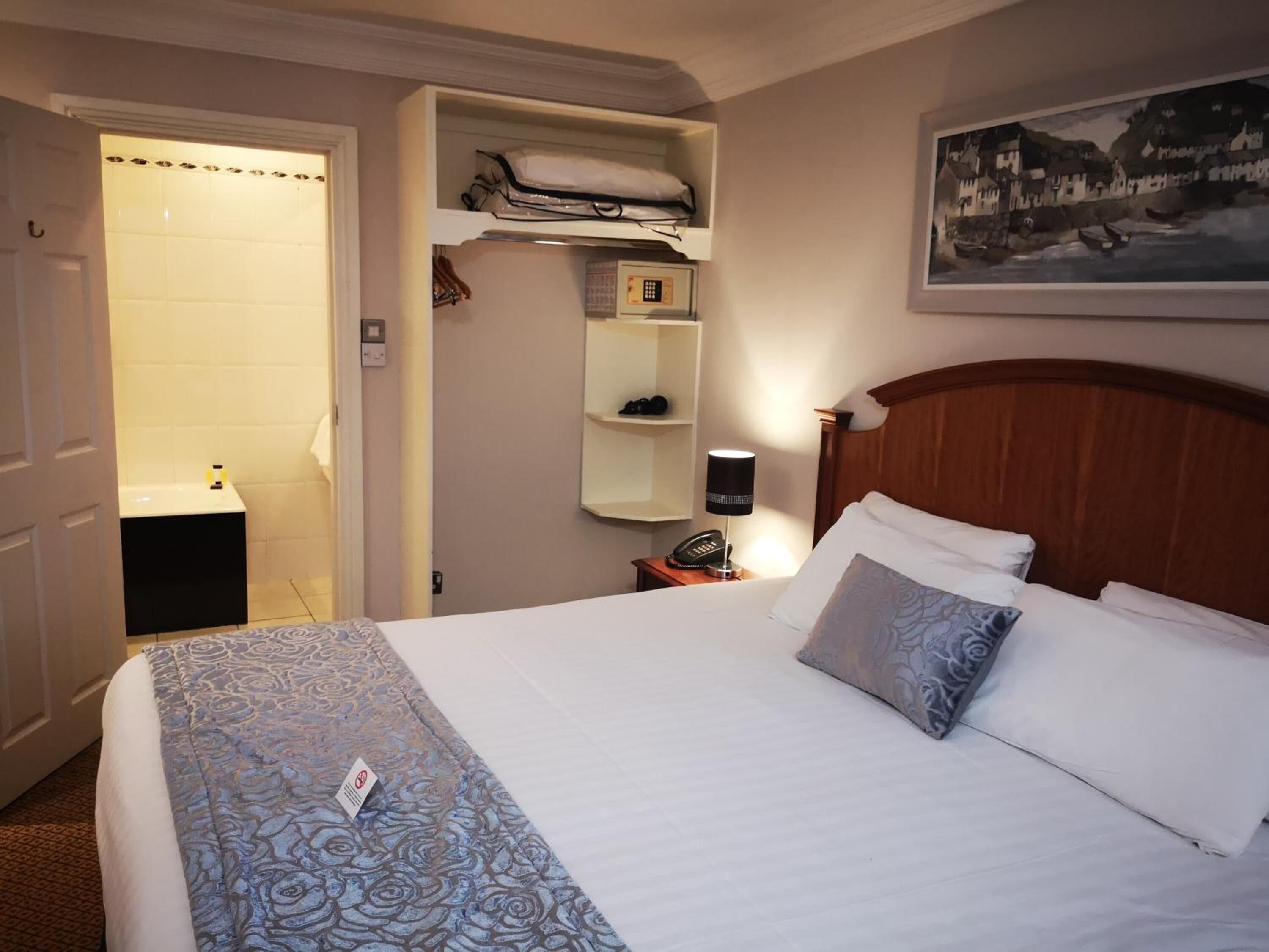 Quarto de hotel com cama, mesa de cabeceira, luminária, pintura na parede e banheiro visível através de uma porta aberta.