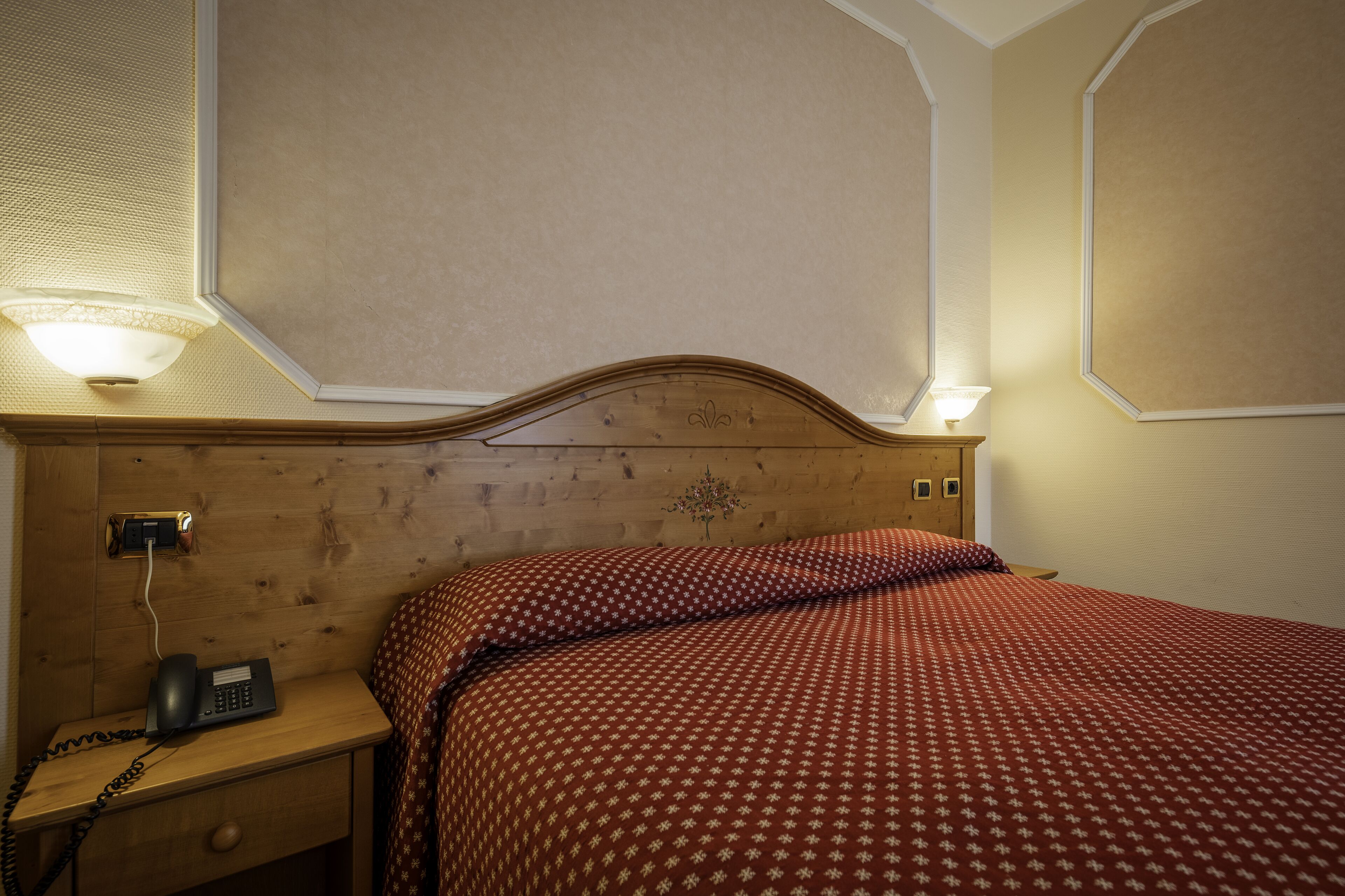 Quarto de hotel com cabeceira de madeira, telefone na mesa de cabeceira e colcha vermelha com padrão.