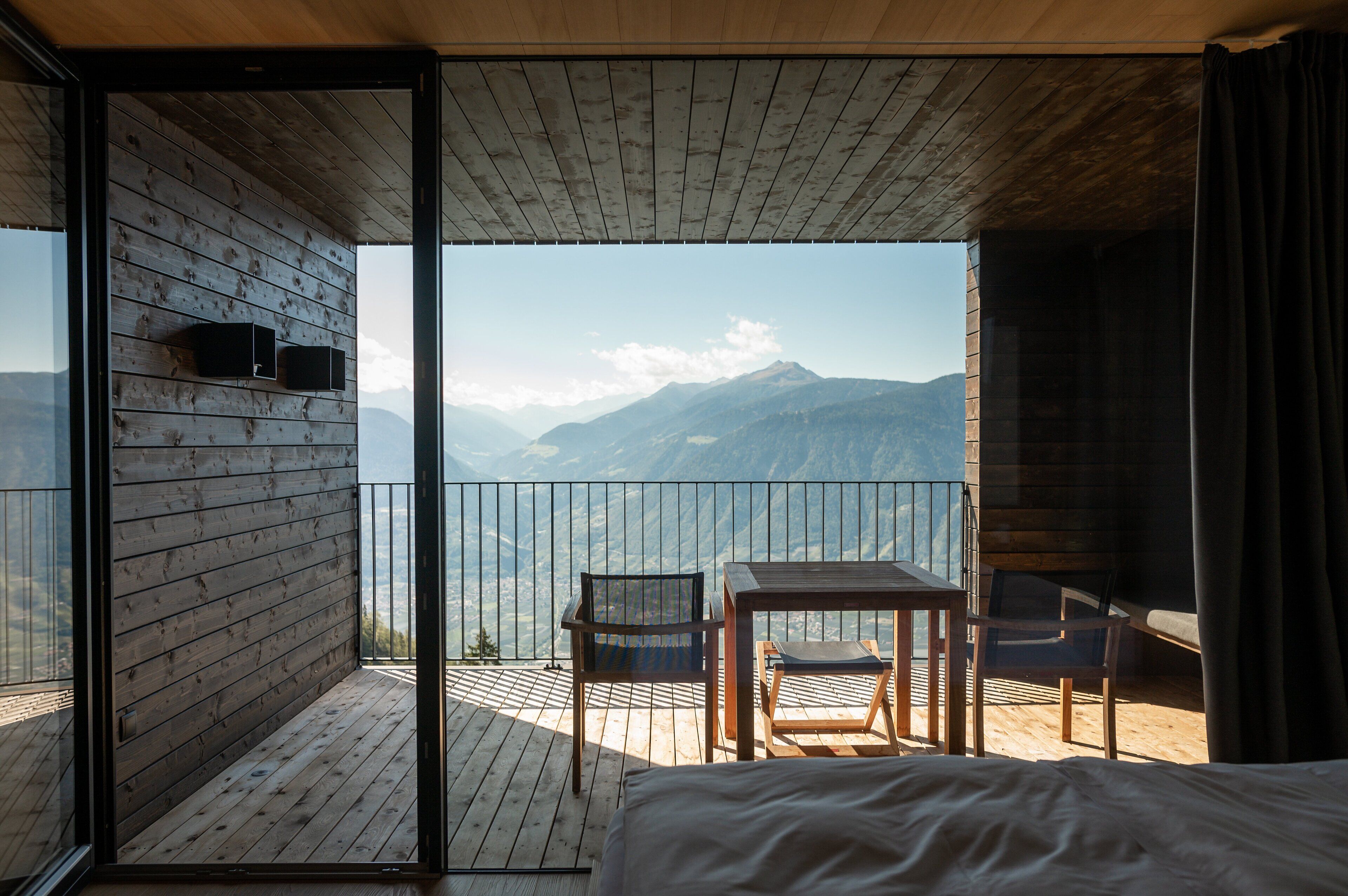 Quarto com piso de madeira, uma cama, uma mesa e duas cadeiras. Possui uma varanda com vista para montanhas.