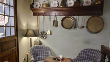 Quarto aconchegante com poltronas xadrez, uma mesa de madeira e uma prateleira fixada na parede com pratos e utensílios de cozinha.