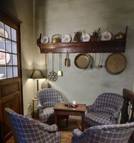 Quarto aconchegante com poltronas xadrez, uma mesa de madeira e uma prateleira fixada na parede com pratos e utensílios de cozinha.