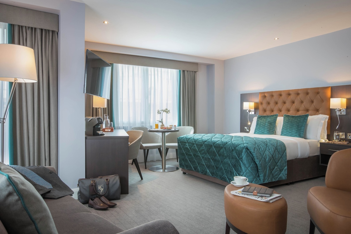 Quarto de hotel com uma cama, uma escrivaninha, uma poltrona, um sofá e uma mesinha de cabeceira com abajur.
