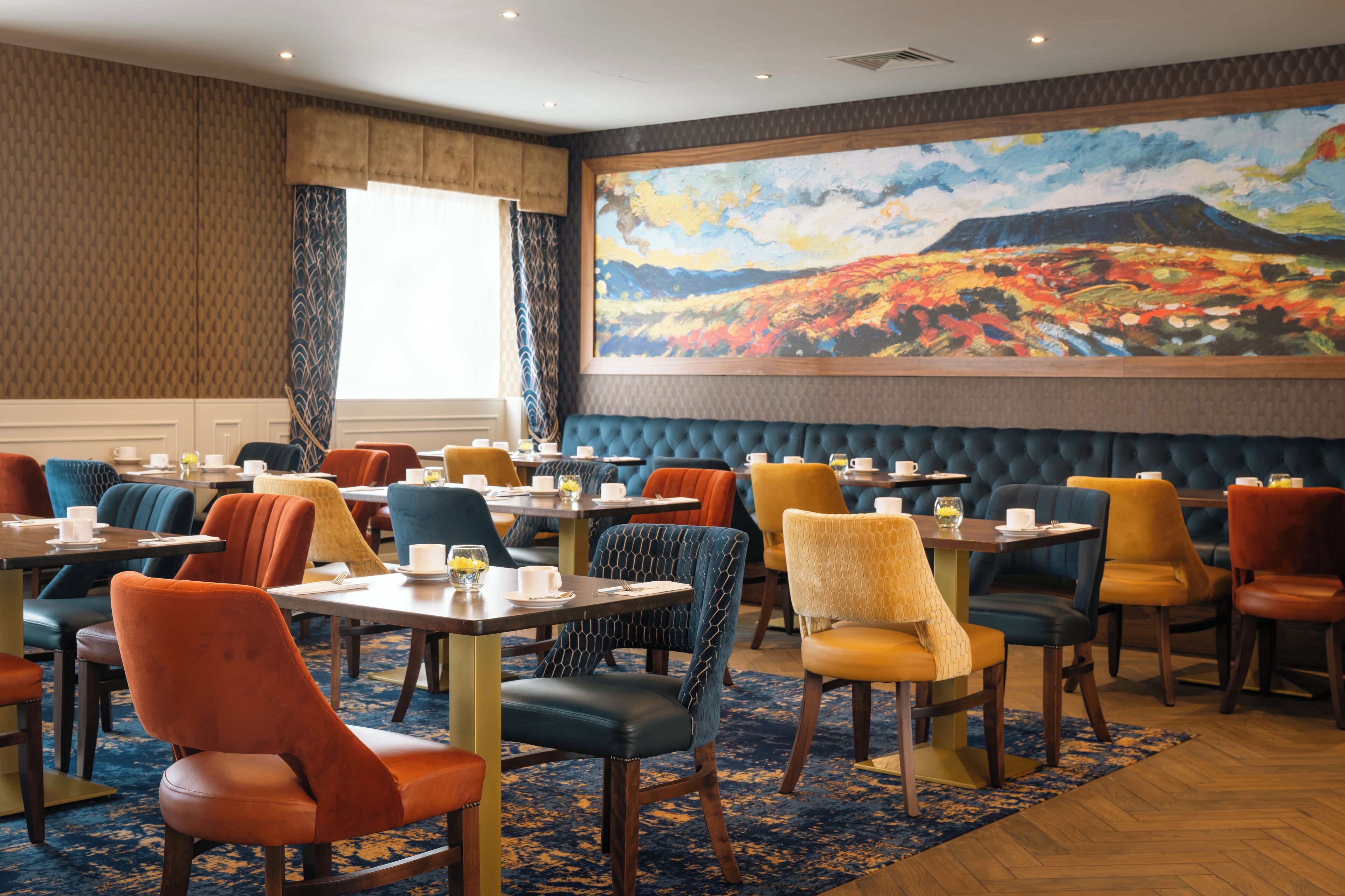 Uma sala de jantar de hotel com várias mesas e cadeiras, um grande mural na parede e um tapete estampado.