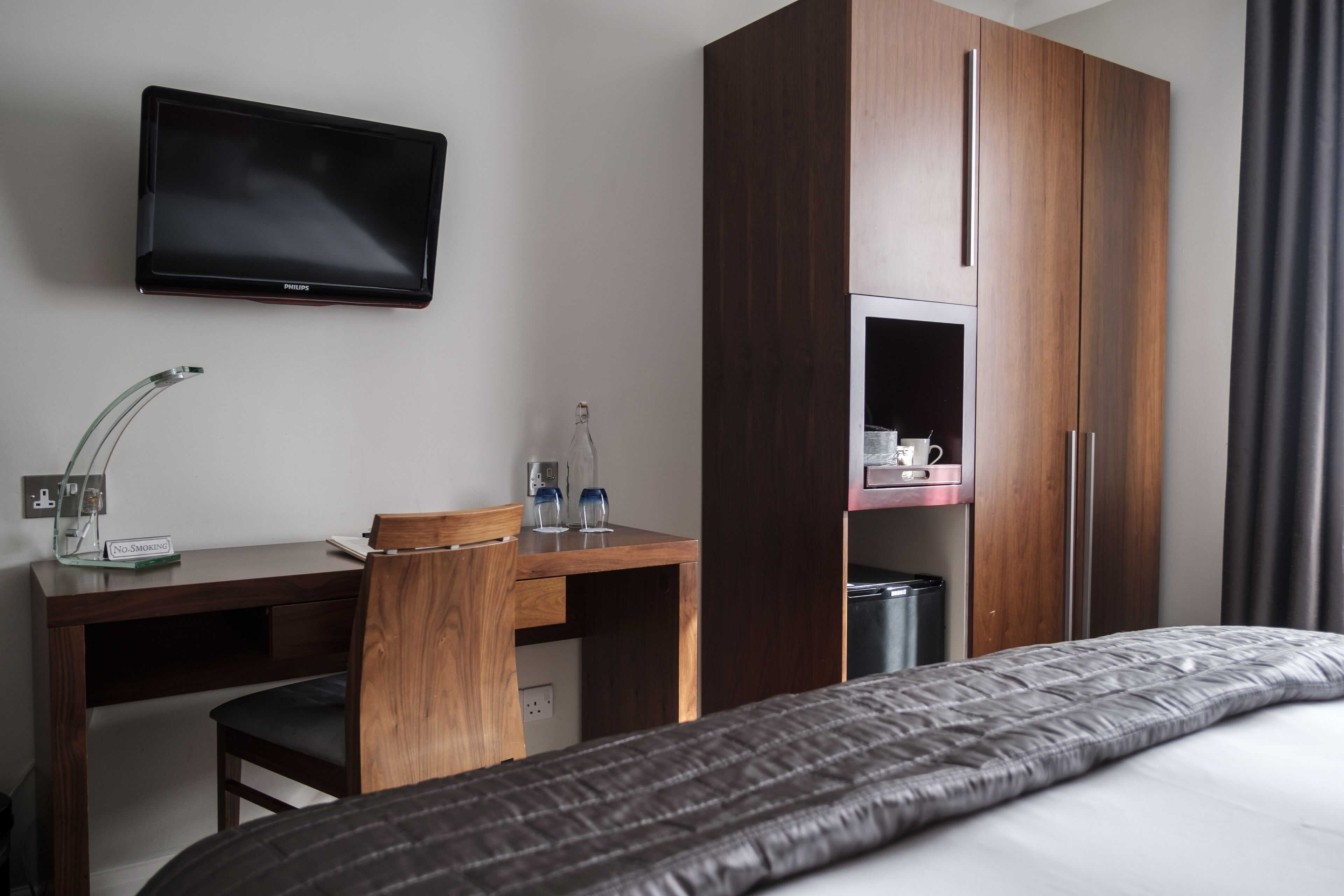 Quarto de hotel com cama, escrivaninha com cadeira, televisão fixada na parede e guarda-roupa.