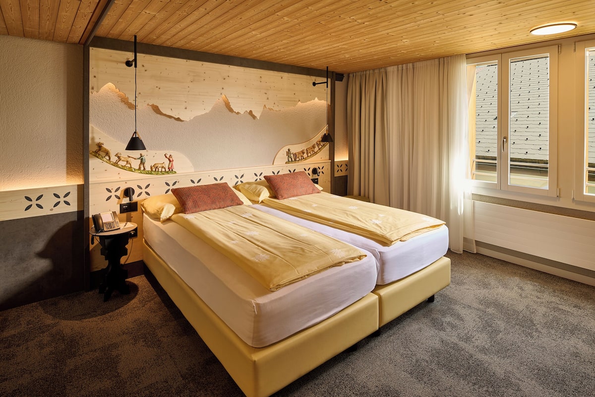 Uma cama de casal com cabeceira decorada com tema de montanha, mesa de cabeceira com abajur e janela com cortinas.