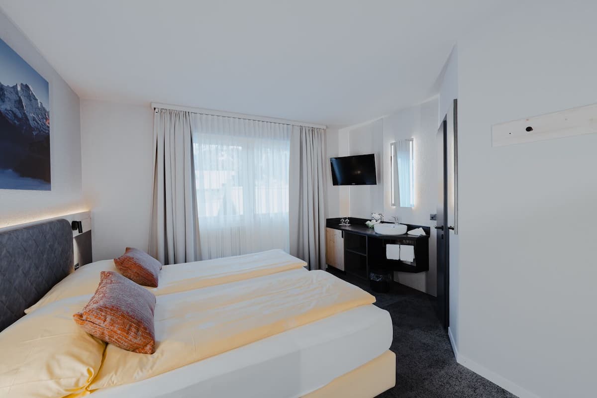 Quarto de hotel com cama, televisão, banheiro com pia e espelho, e janela com cortinas.