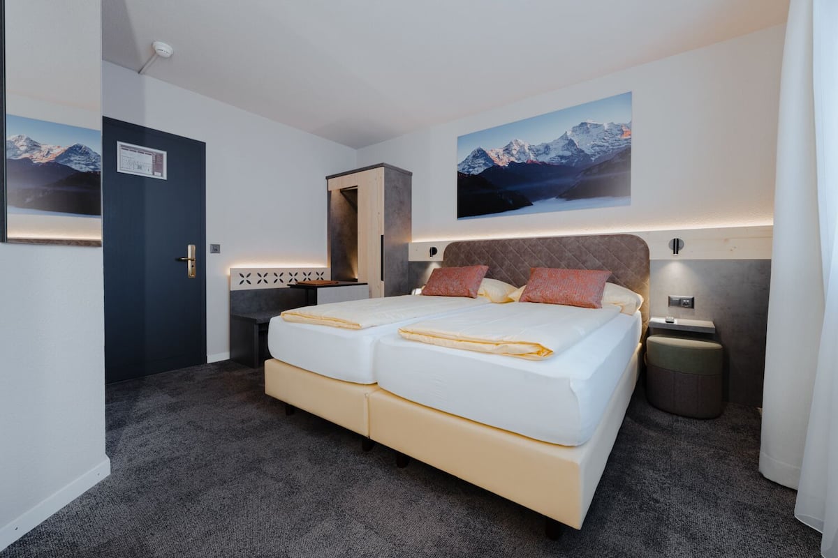 Uma cama de casal com cabeceira, mesa de cabeceira, lixeira e painel de controle fixado na parede.