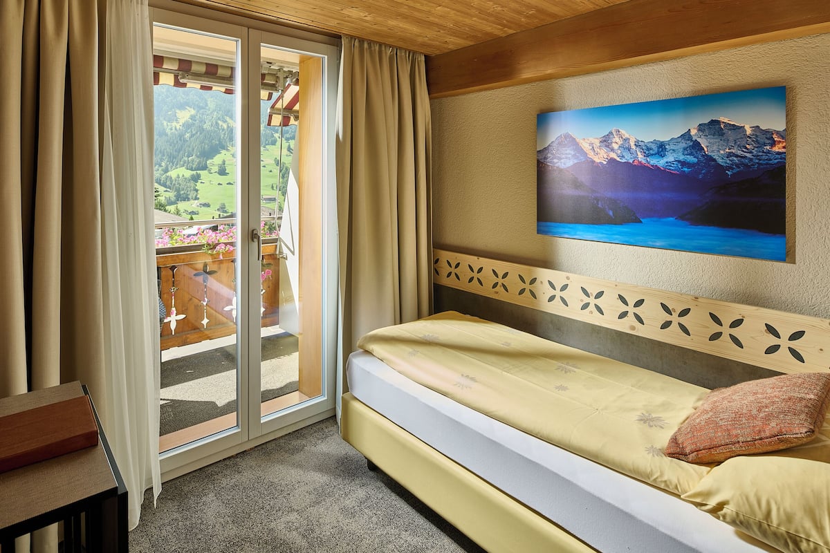Quarto com cama, janela com cortinas, pintura de montanhas e varanda florida.