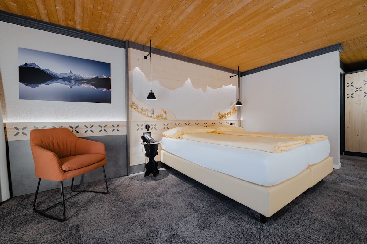 Quarto com cama, cadeira, luminária na parede e pintura de montanhas.