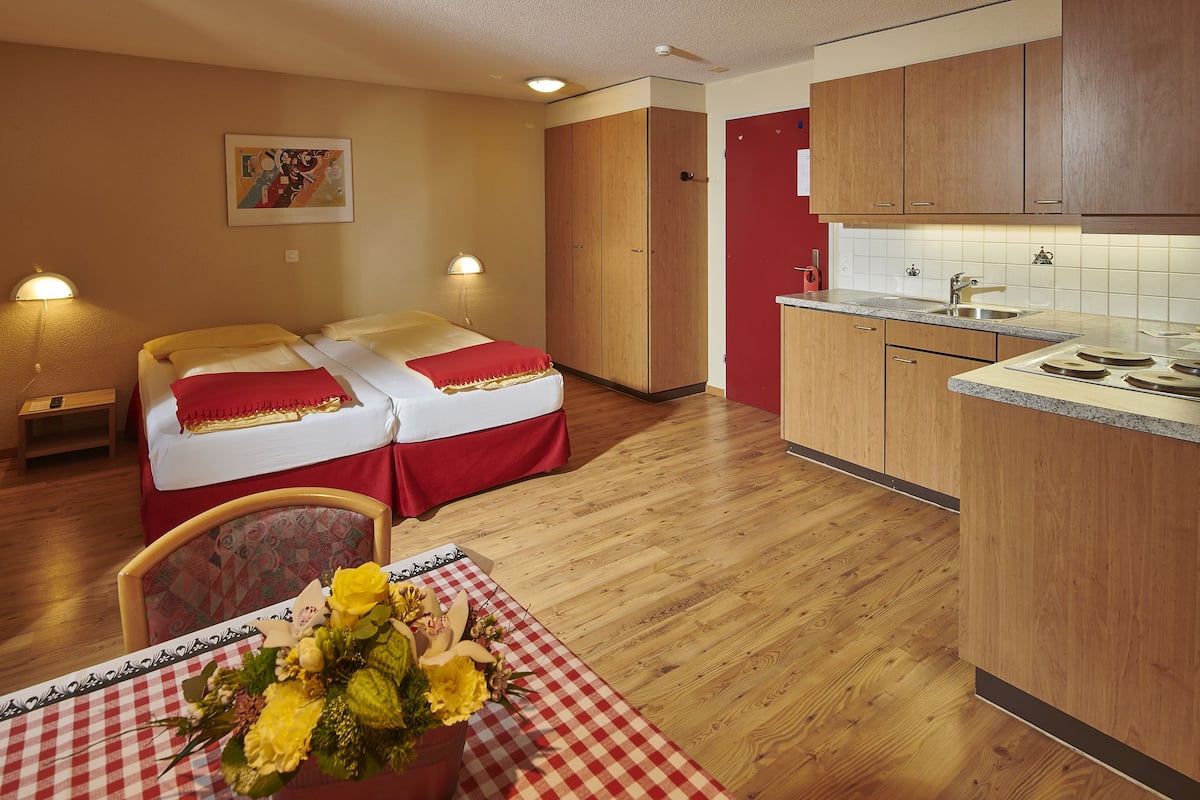 Quarto de hotel pequeno com duas camas, área de refeições com mesa xadrez e cozinha compacta.