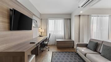 Quarto de hotel moderno com escrivaninha de madeira, televisão de tela plana, sofá e uma janela grande com cortinas leves.
