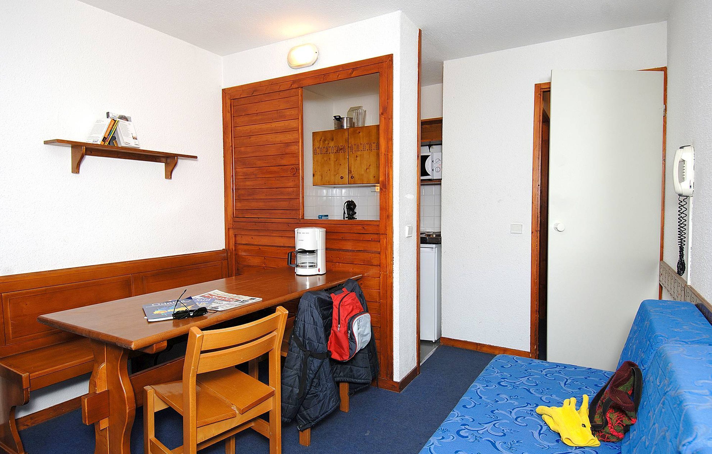 Quarto compacto com mesa de madeira, uma cadeira, cama com colcha azul, estante com livros e pequena área de cozinha.