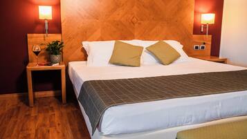 Quarto de hotel com cabeceira de madeira, cama com lençóis brancos e duas almofadas, mesa de cabeceira com abajur, uma pequena planta e uma taça de vinho.