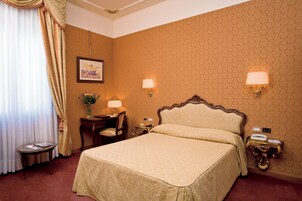 Quarto de hotel com uma cama grande, duas mesas de cabeceira, uma escrivaninha com cadeira, um abajur e um quadro na parede.