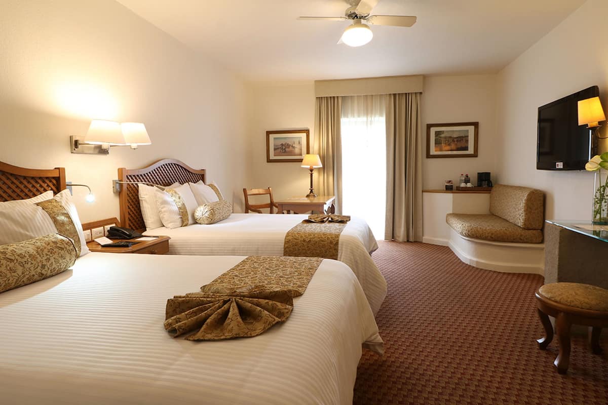 Quarto de hotel com duas camas, uma escrivaninha, uma cadeira, uma televisão e um ventilador de teto.