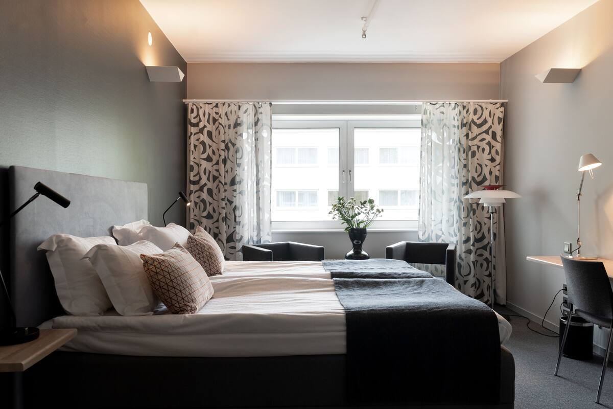 Quarto de hotel moderno com uma cama grande, mesas de cabeceira, escrivaninha e janela com cortinas estampadas.