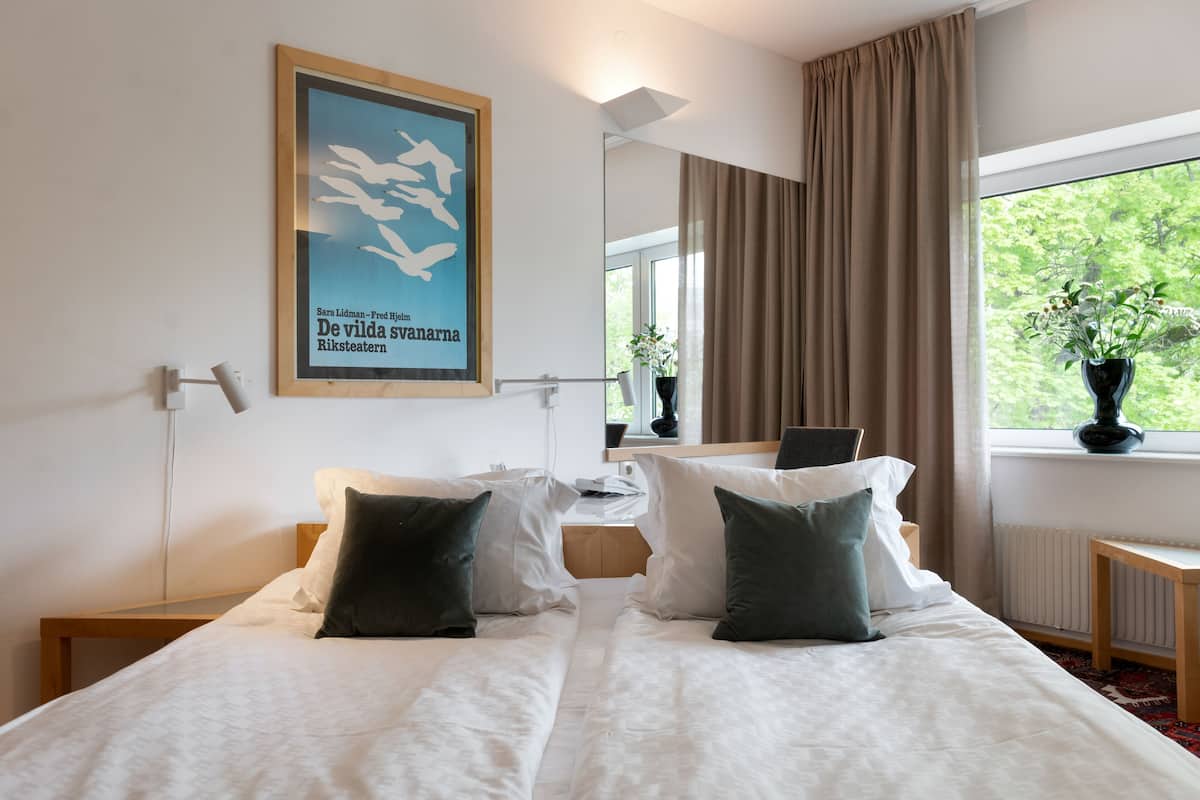 Quarto de hotel com cama, janela com cortinas, mesa de cabeceira, luminária e quadro na parede.