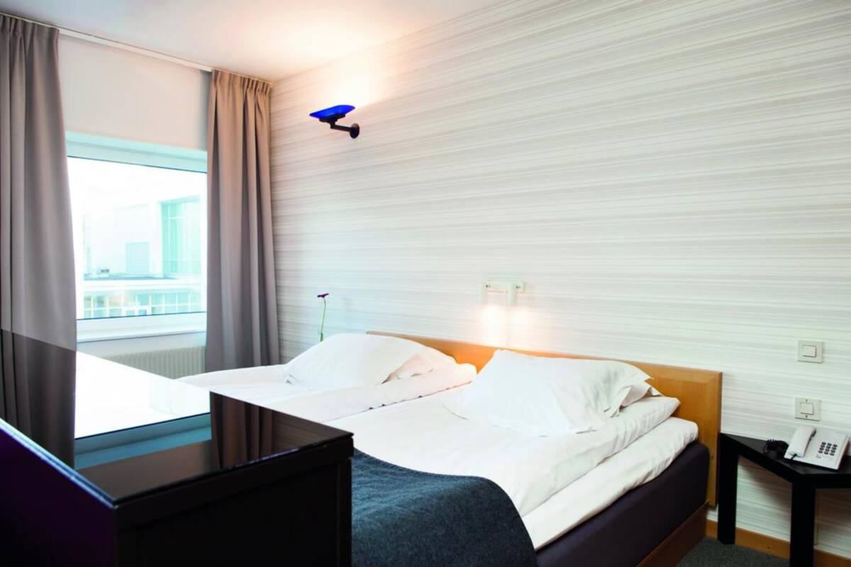 Quarto de hotel com cama, escrivaninha, telefone e janela com cortinas.