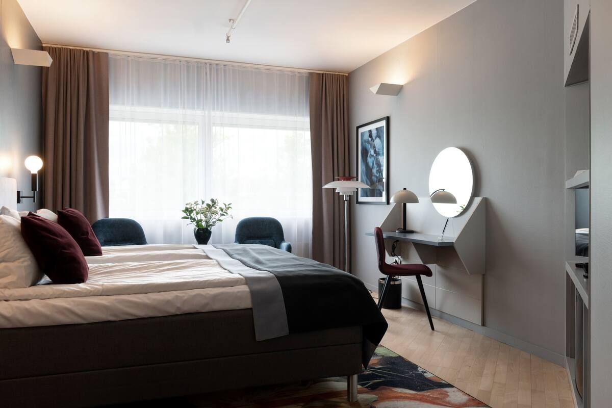 Quarto de hotel moderno com uma cama grande, duas cadeiras, escrivaninha com computador, luminária de mesa, espelho redondo e janela com cortinas leves.