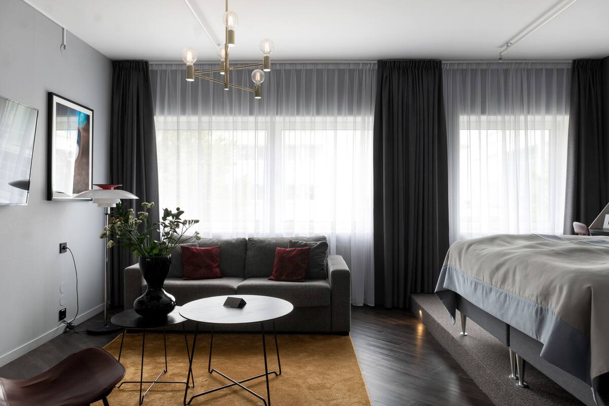 Quarto de hotel moderno com uma cama grande, um sofá, uma mesa de centro redonda e uma planta em vaso.