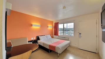 Quarto de hotel com cama, escrivaninha, cadeira, janela com cortinas e porta.