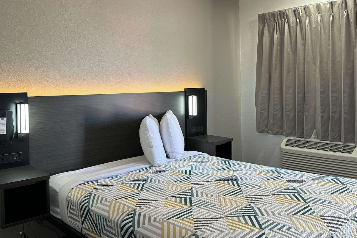 Quarto de hotel com cama, duas mesas de cabeceira com abajures, ar-condicionado na parede e janela com cortinas.