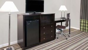 Quarto de hotel com um armário de madeira escura, uma TV de tela plana, um frigorífico, uma secretária com cadeira, duas luminárias e um tapete riscado.