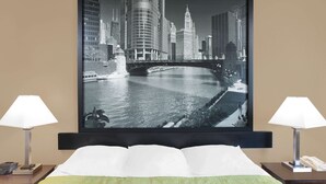 Quarto de hotel com um mural grande em preto e branco da paisagem urbana na parede, uma cama com roupa de cama branca e verde, duas luminárias de cabeceira e uma mesa de cabeceira com um telefone e um controle remoto.