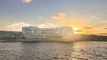 Hotel à beira-mar com vários andares e varandas, localizado perto da água onde o pôr do sol se reflete.