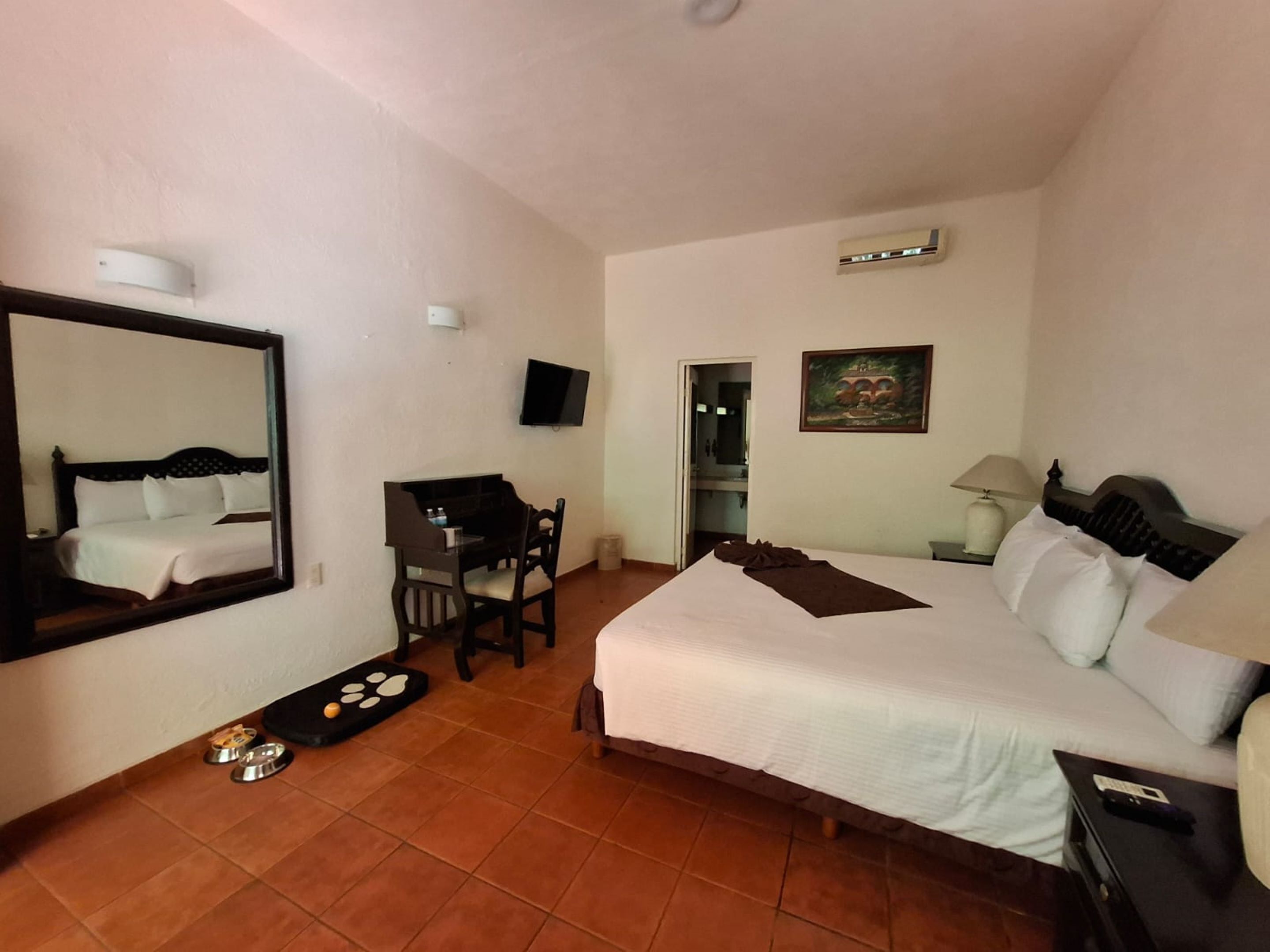 Quarto de hotel com cama, escrivaninha, cadeira, televisão, um quadro e banheiro visível através de uma porta aberta.