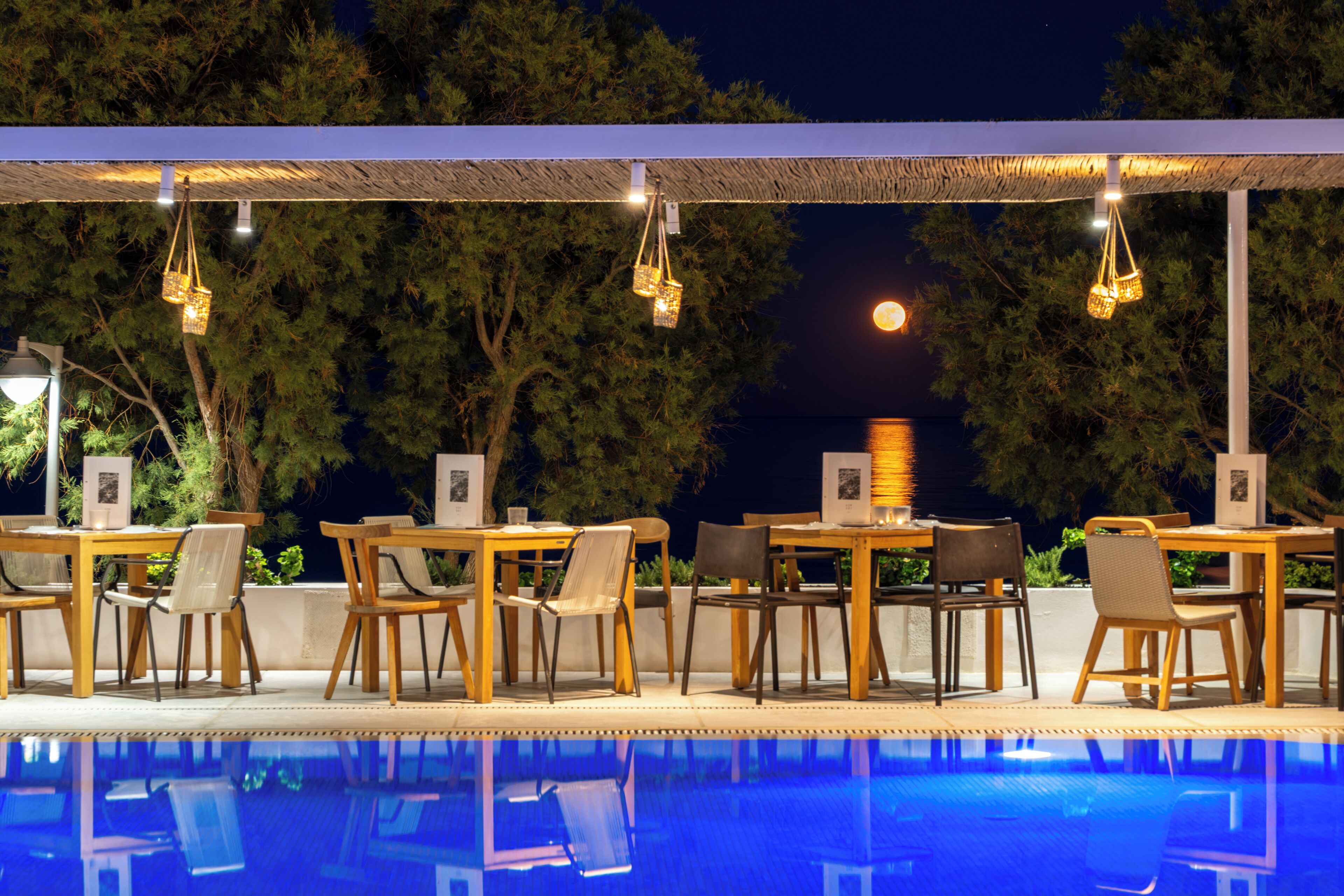 Uma área de piscina ao ar livre com mesas e cadeiras, rodeada por árvores e um céu noturno.