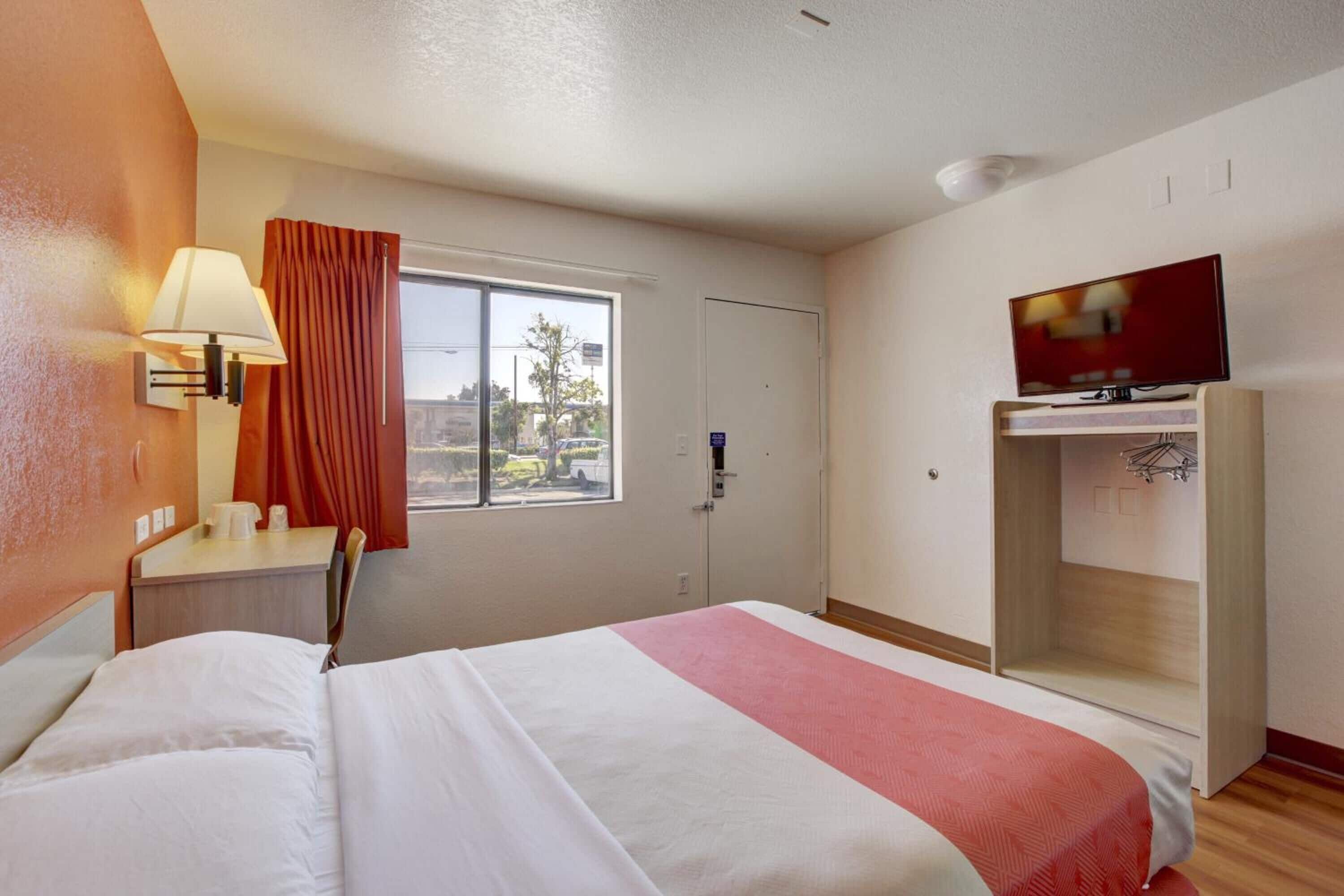 Quarto de hotel com cama, televisão em móvel, janela com cortinas e escrivaninha com abajur.