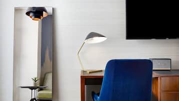 Sala de estar moderna com televisão de tela plana, uma poltrona azul, escrivaninha com laptop, luminária de chão e mesa lateral com planta.