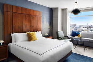 Quarto de hotel com uma cama grande, vista para a cidade, uma cadeira moderna e uma mesinha.