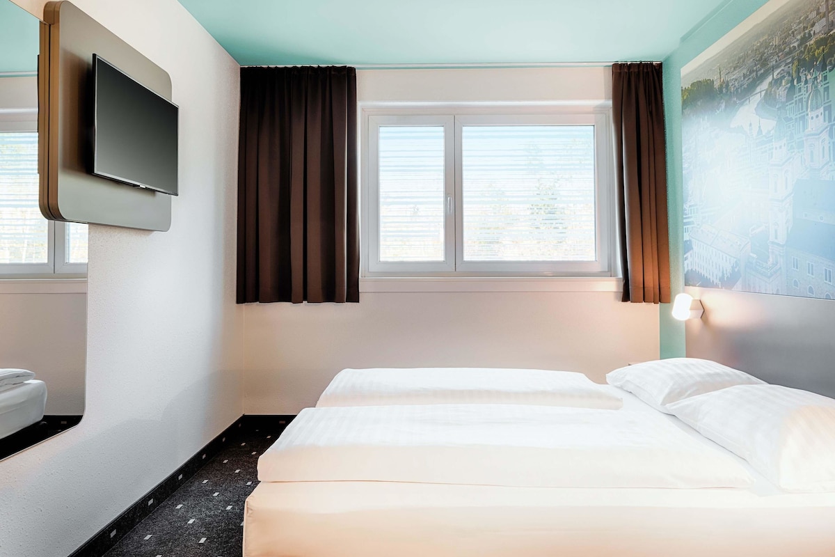 Quarto de hotel com duas camas, uma TV de tela plana fixada na parede, uma janela com cortinas e um quadro na parede.