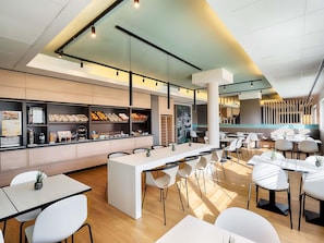 Um café moderno com um balcão central exibindo pastelaria e bebidas, rodeado por mesas e cadeiras brancas.