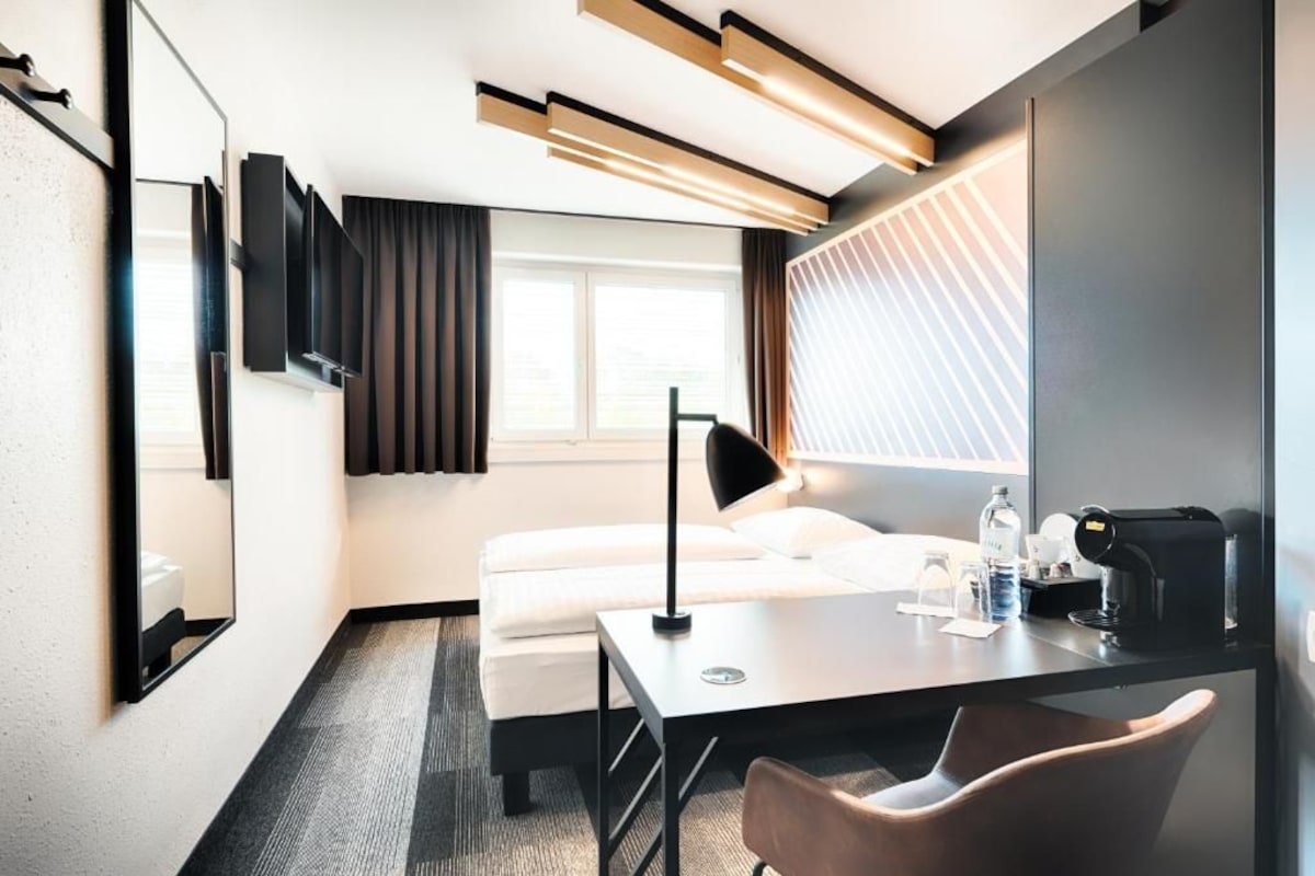 Quarto de hotel moderno com cama, escrivaninha, cadeira e janela com persianas.