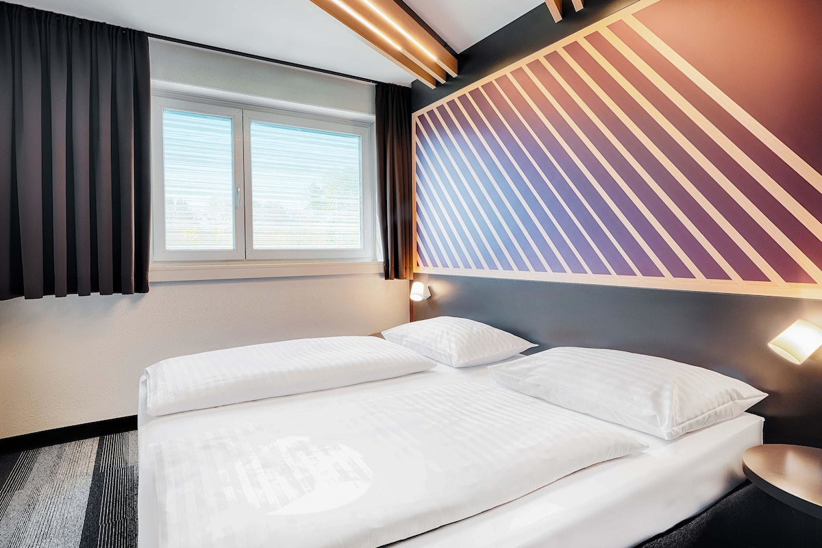Quarto de hotel com cama, janela com persianas, parede com padrão listrado e mesa de cabeceira com abajur.
