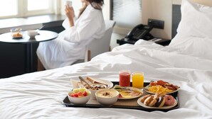 Quarto de hotel com cama, bandeja de café da manhã e uma pessoa usando o celular.